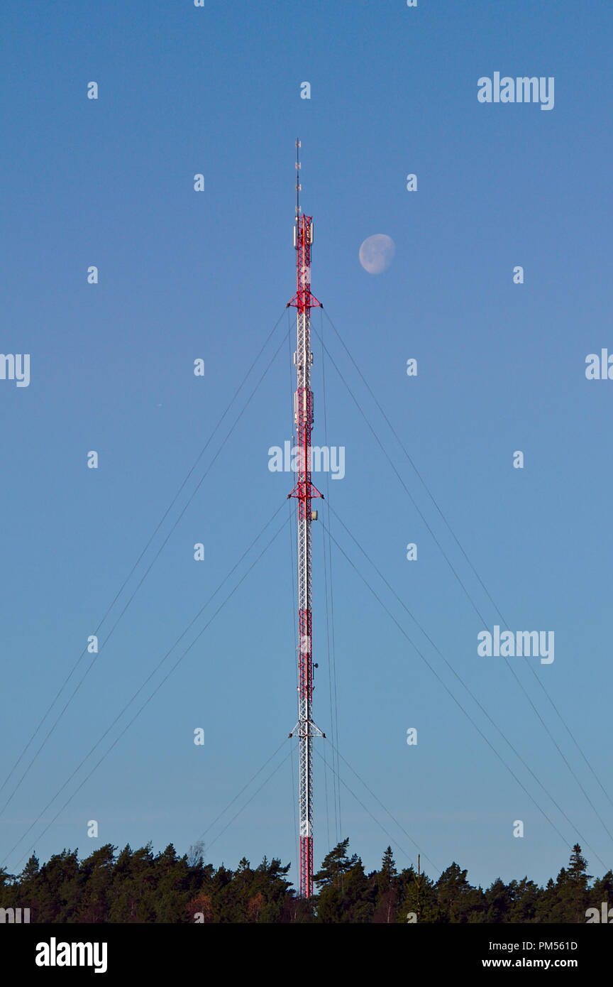 High red and white cellular tower at daytime with moon shining next to the top of the tower ...