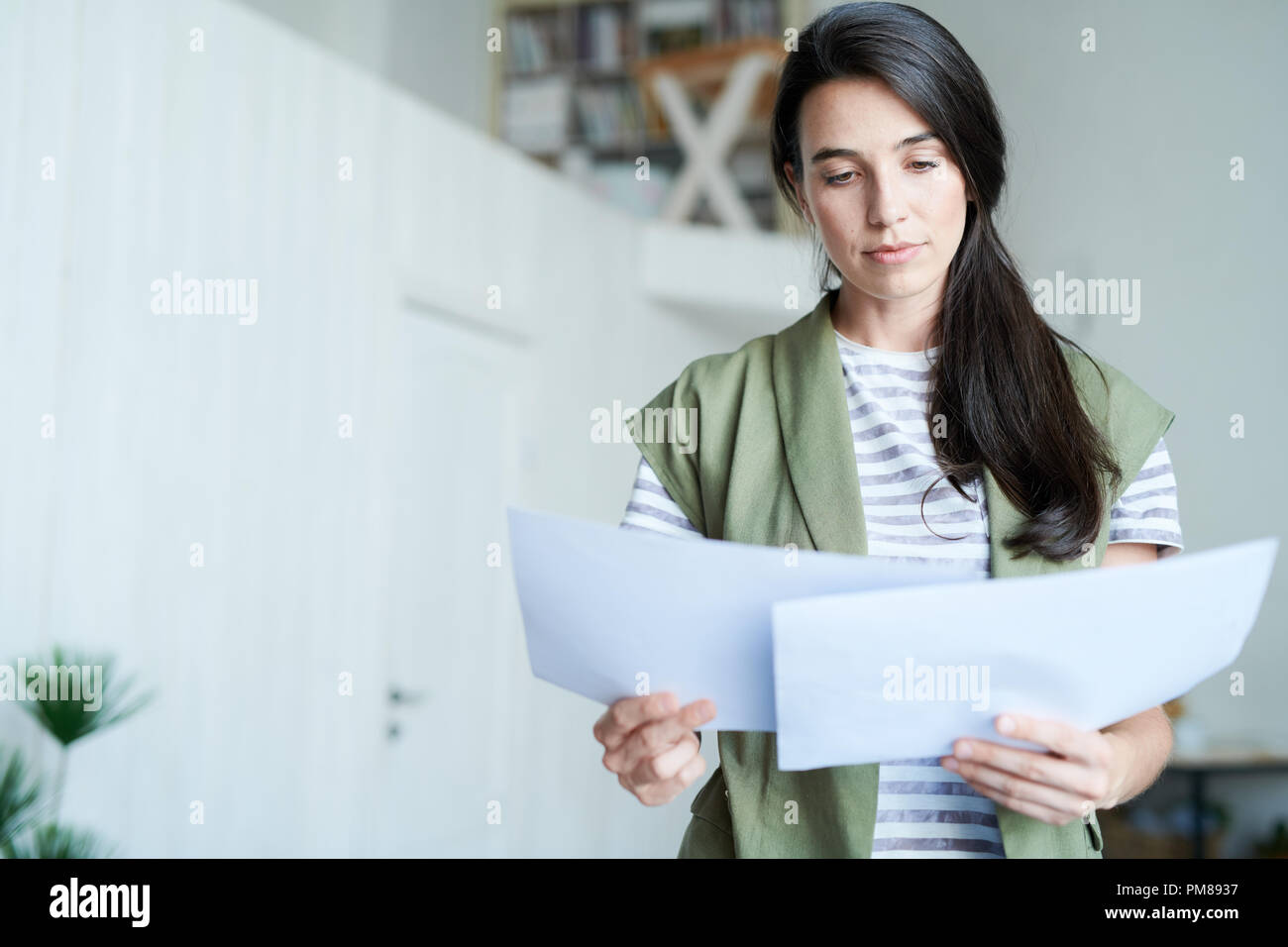 Portrait of brunette young woman holding two documents and reading them ...