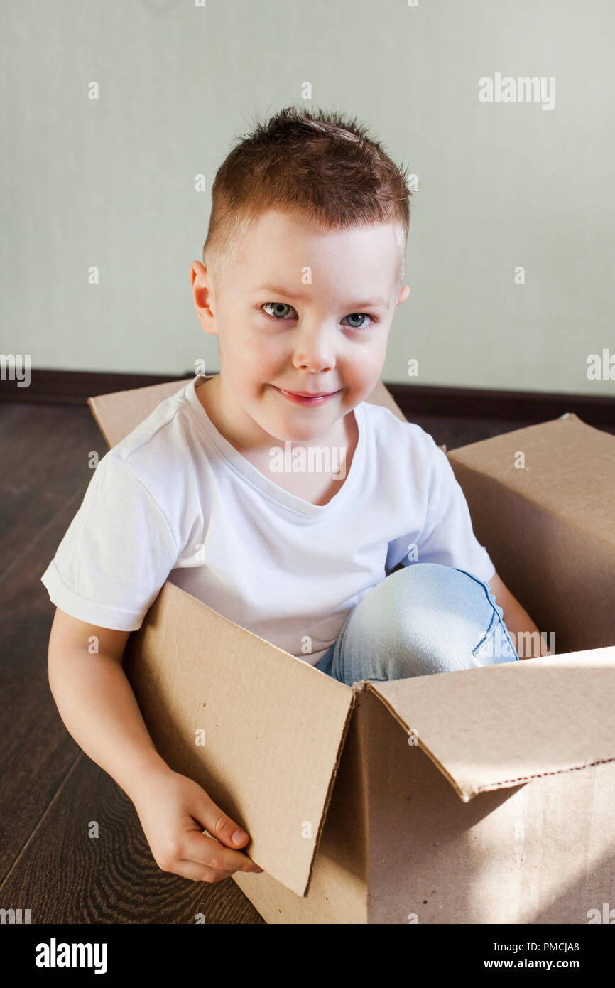 A 4 Year Old Blonde Boy Sits In A Box At Home And Looks At The Camera