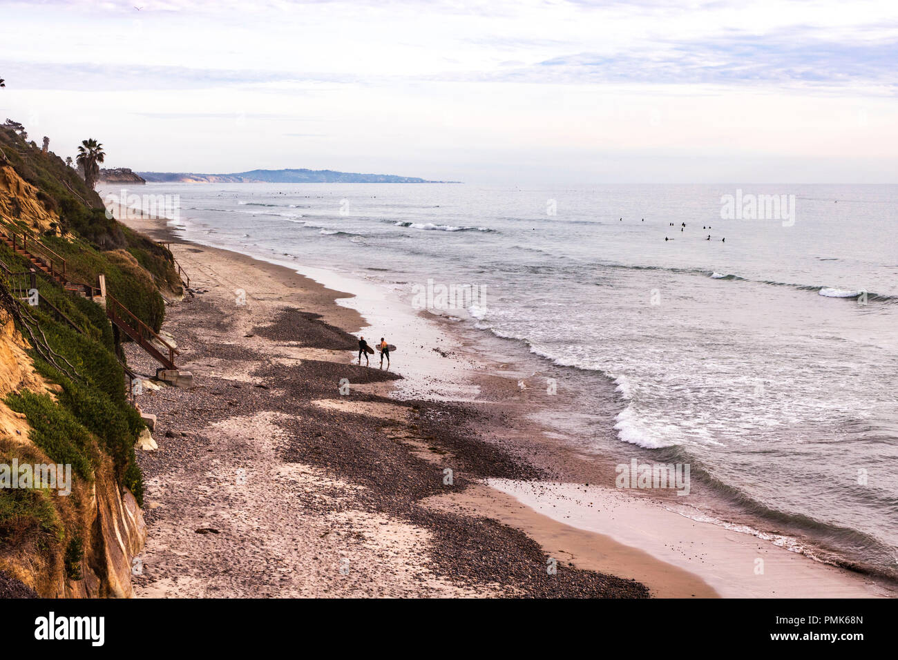 Two surfers walk up a rocky shoreline beach with surf rolling in, green ...