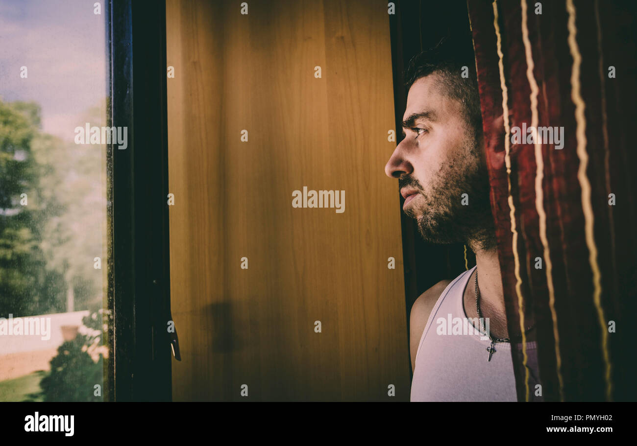 Thoughtful young man among the curtains looking out the window of his ...