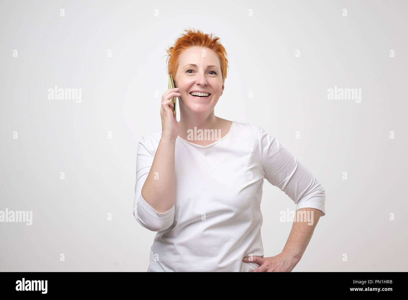 Smiling confident woman with short red hairstyle talking on cell phone over white background ...