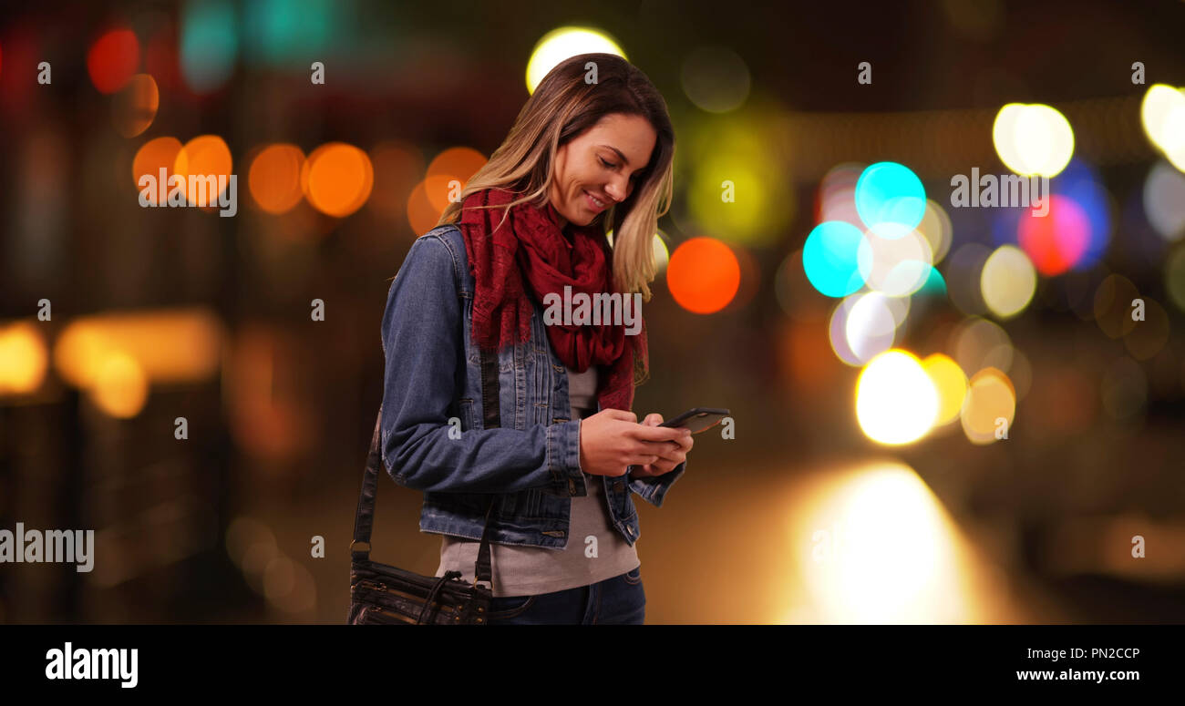 Trendy millennial girl messaging on smartphone on city street at night ...