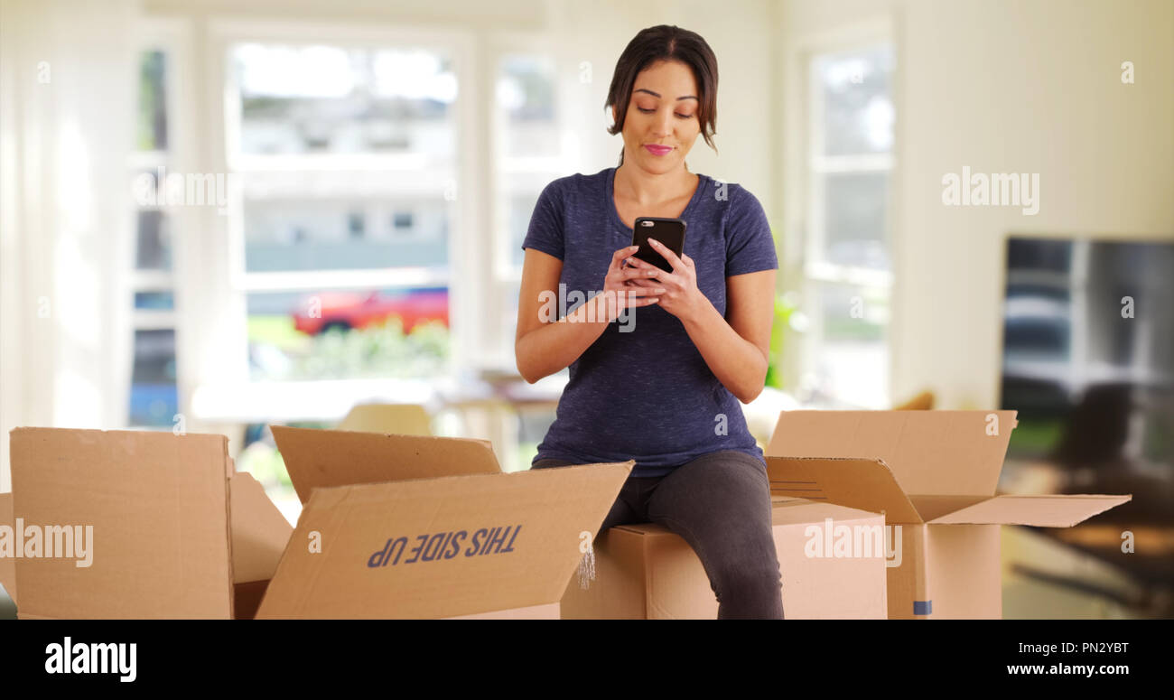 Tired young Latina woman taking break from packing boxes texting on ...