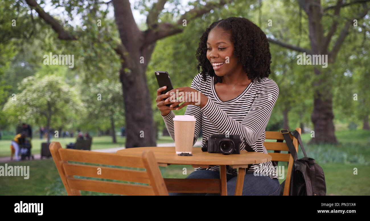 Cute black woman sits at table texting on smartphone at St James park ...