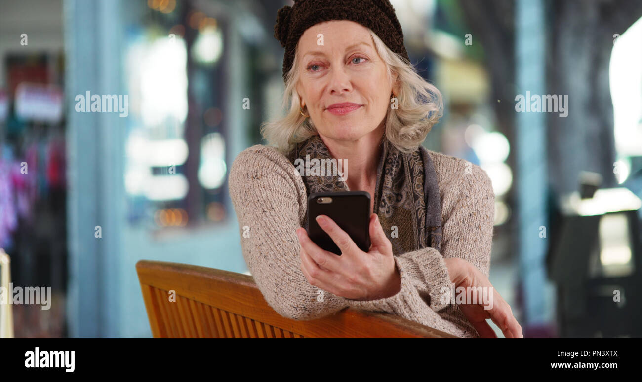 Female senior citizen with smartphone sitting on public bench on city ...