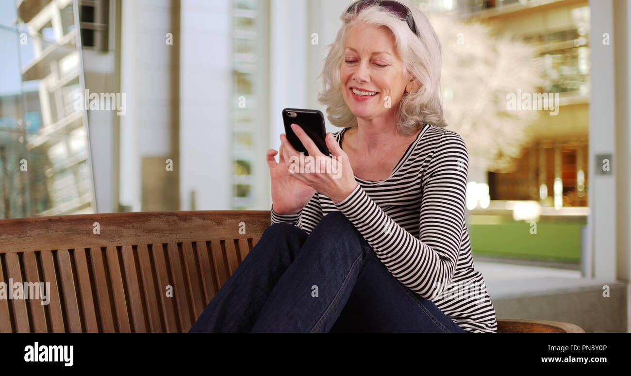 Caucasian woman senior using mobile device sitting on bench by modern ...