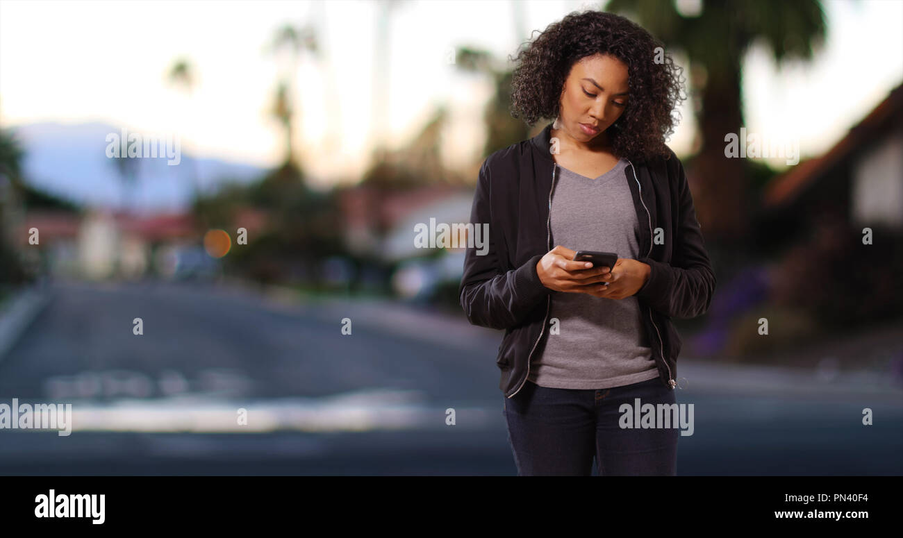 Young black woman sending message on smartphone in residential street ...