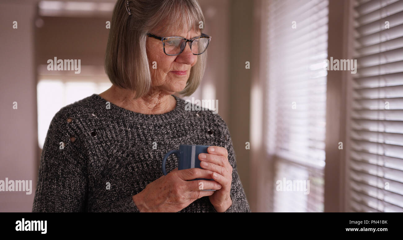 Somber old white woman contemplating and holding cup while looking out ...