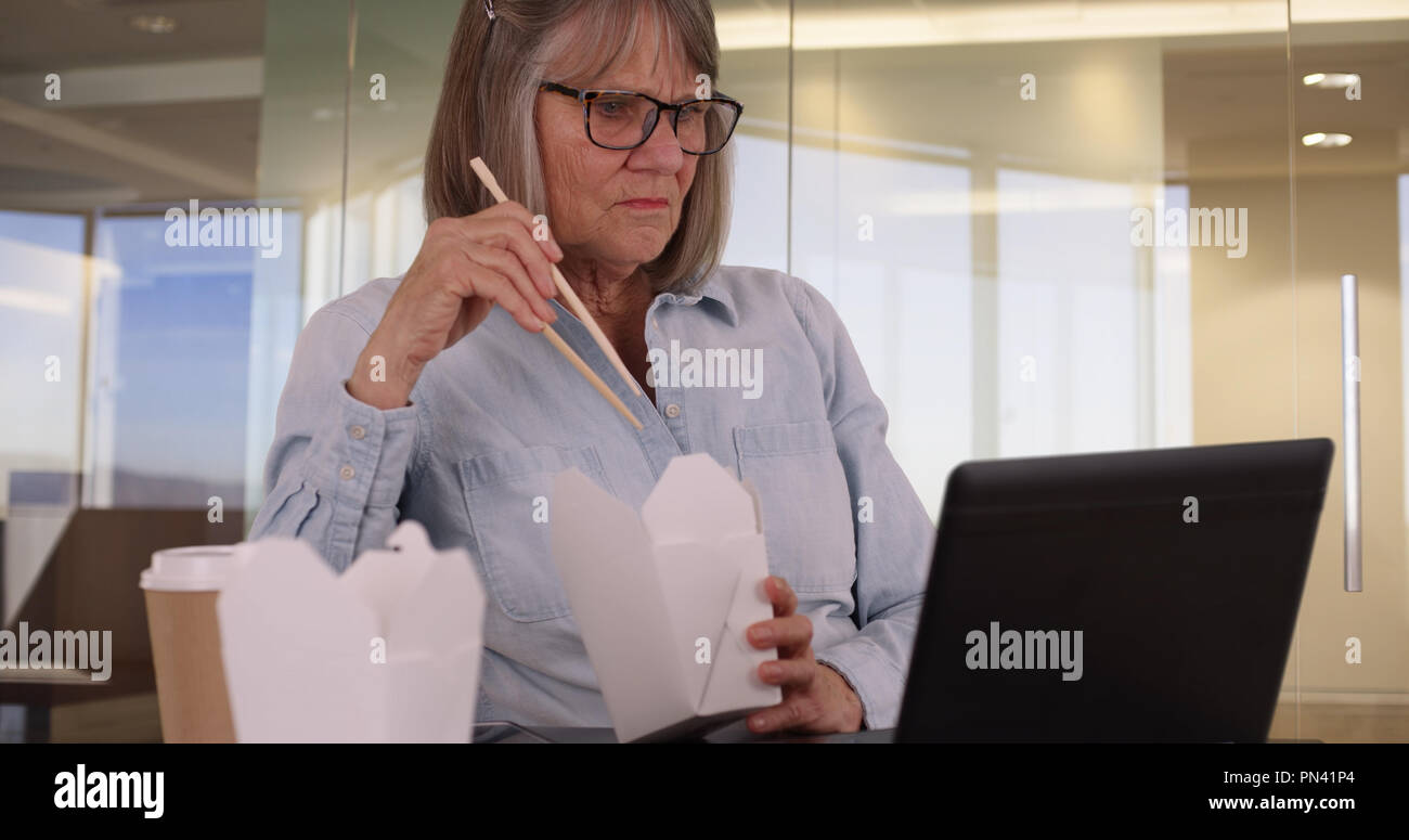 White female corporate employee eating while using laptop in office ...