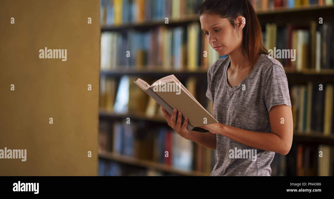 Beautiful young Caucasian woman reading in library between the stacks ...