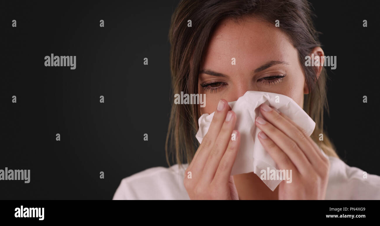 Sick Caucasian woman blowing nose into tissue paper on dark gray ...