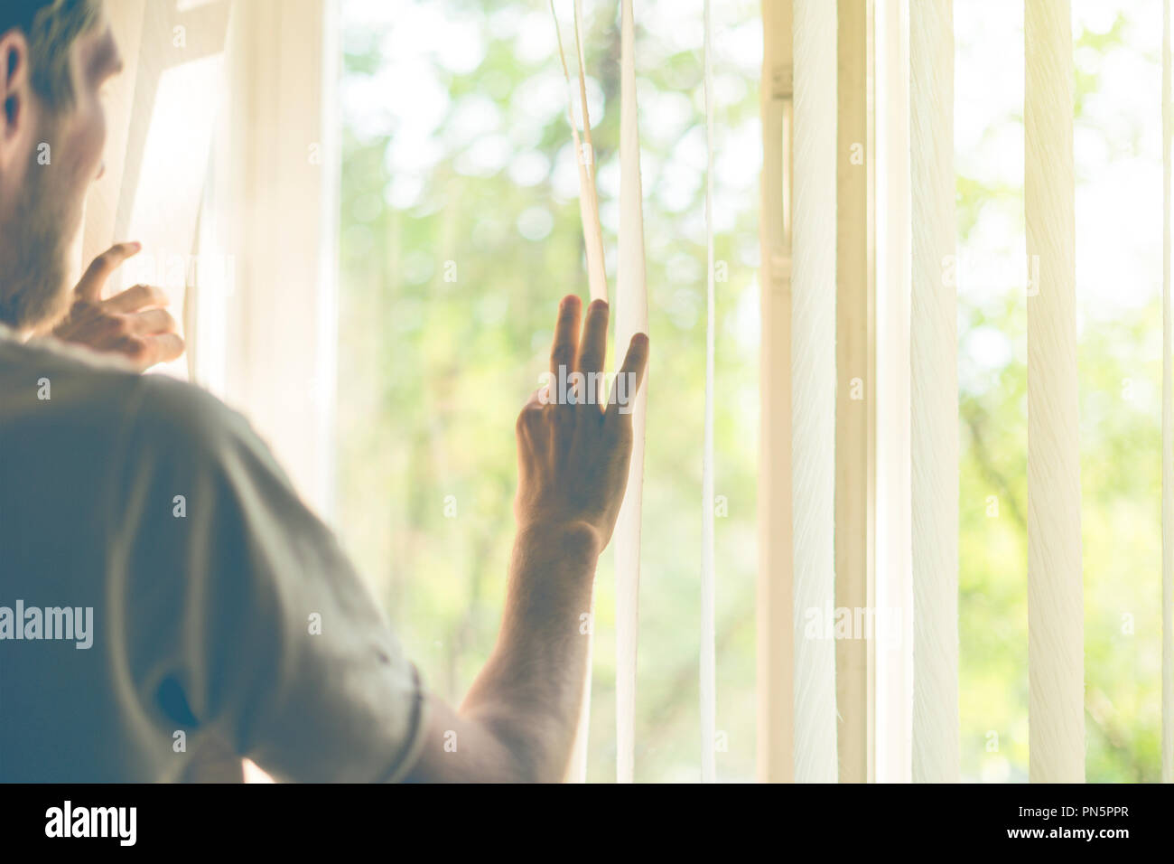back view of man looking through jalousie blinds on widnow at the sun ...