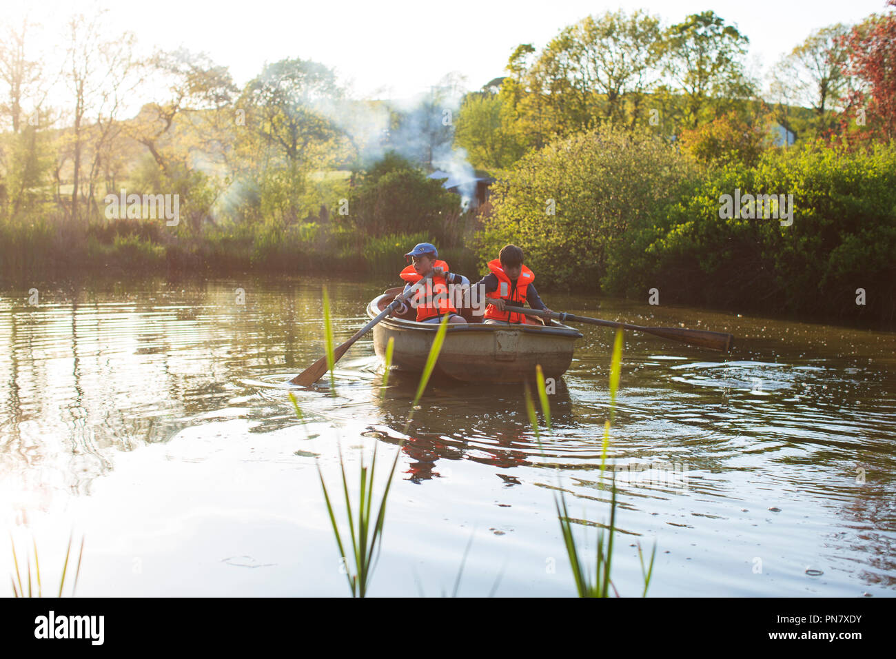 Two boys rowing a boat on a small lake back lit by the evening sun ...