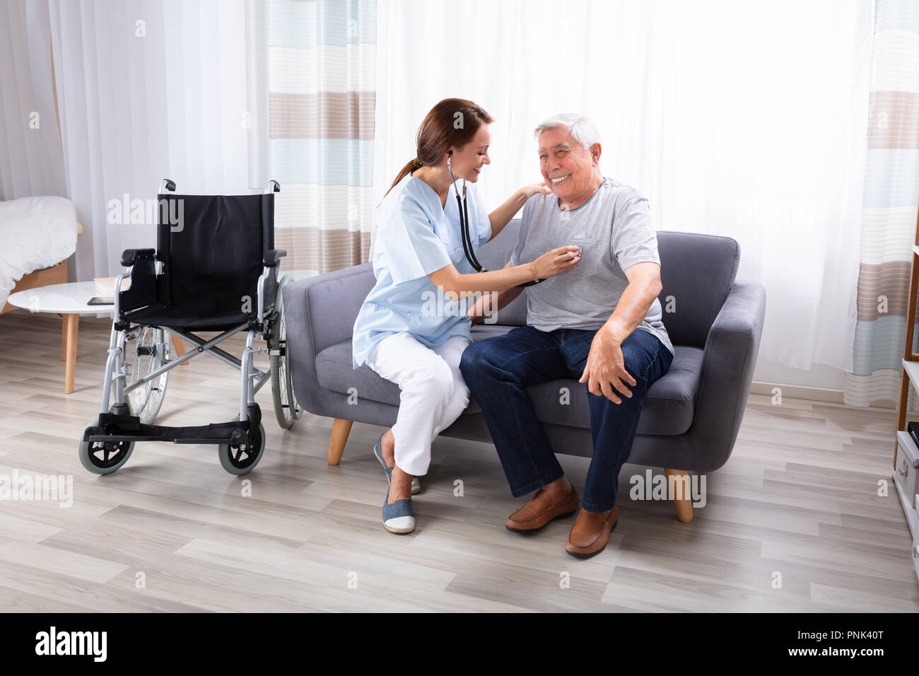 Female Nurse Checking Happy Senior Man Sitting On Sofa With Stethoscope ...