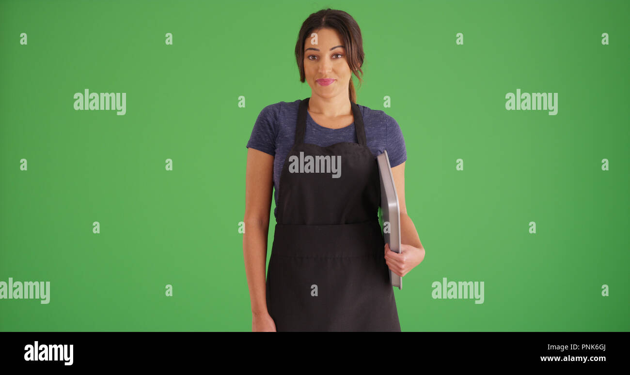 Smiling Hispanic woman wearing apron posing with baking sheet on green ...
