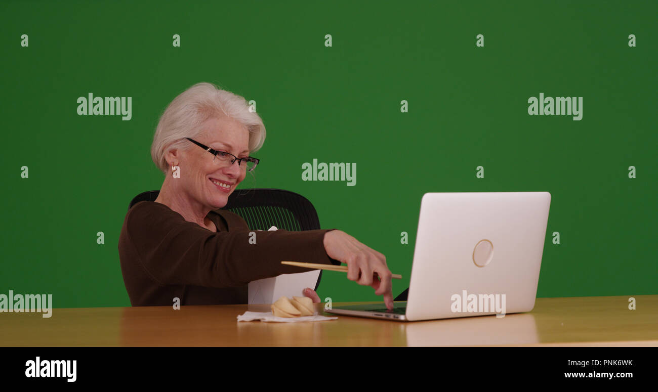 Old business female eating takeout on lunch break using laptop on green ...
