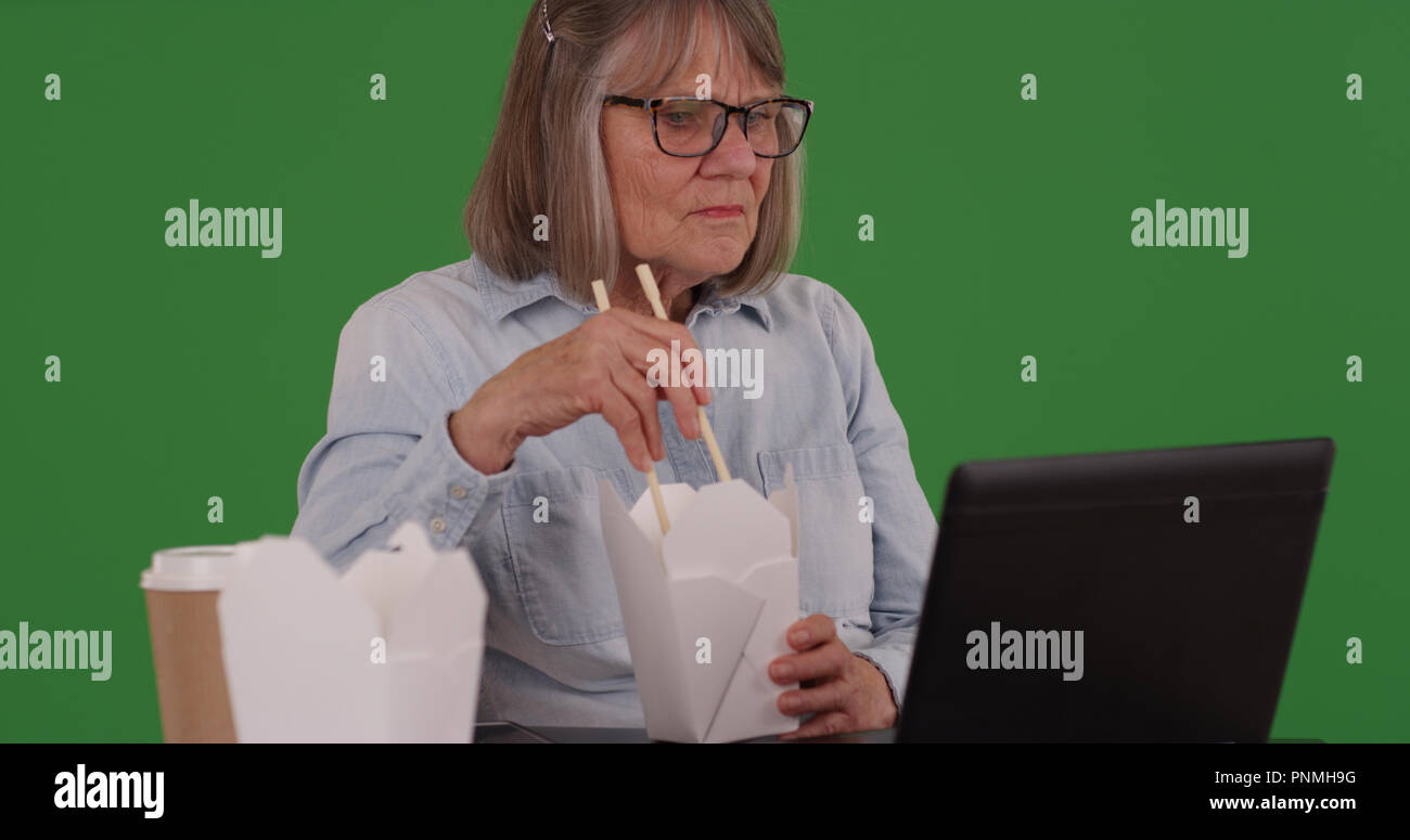 Portrait of senior female eating Chinese food using laptop on green ...