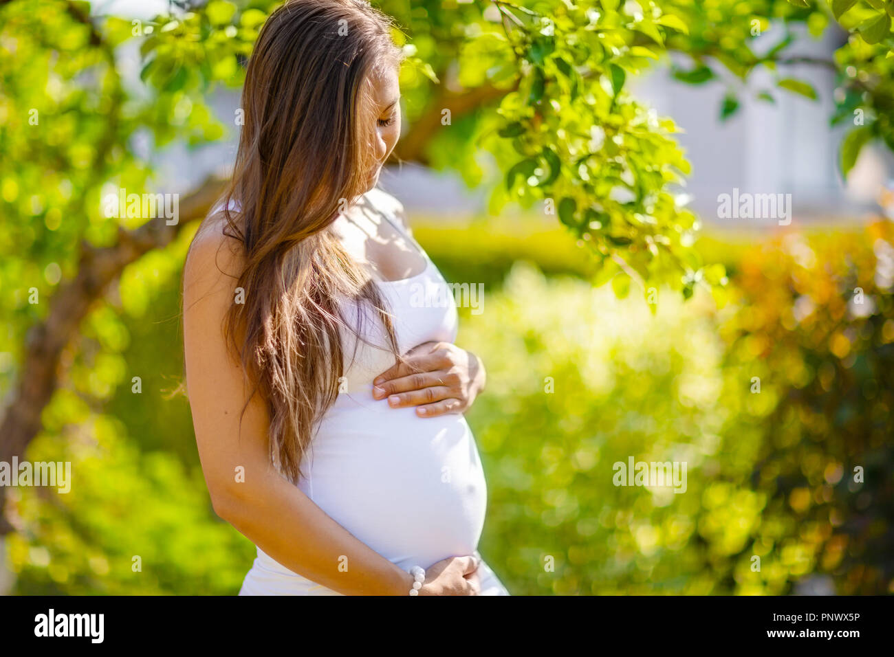 Smiling Pregnant Woman Standing In Garden Holding Hands On Belly Stock