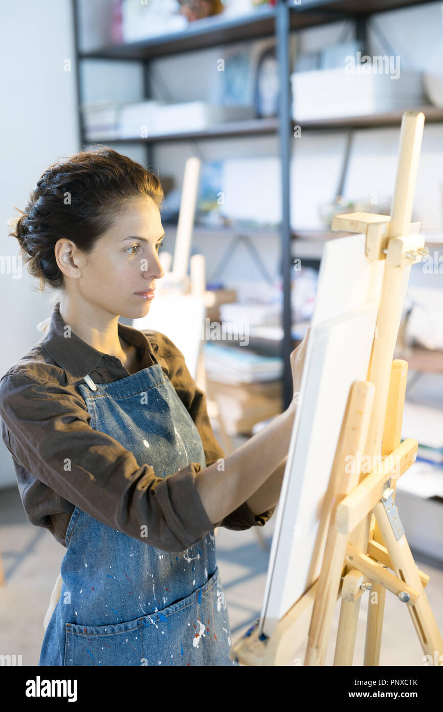 Young female painter standing in front of easel and painting picture of ...