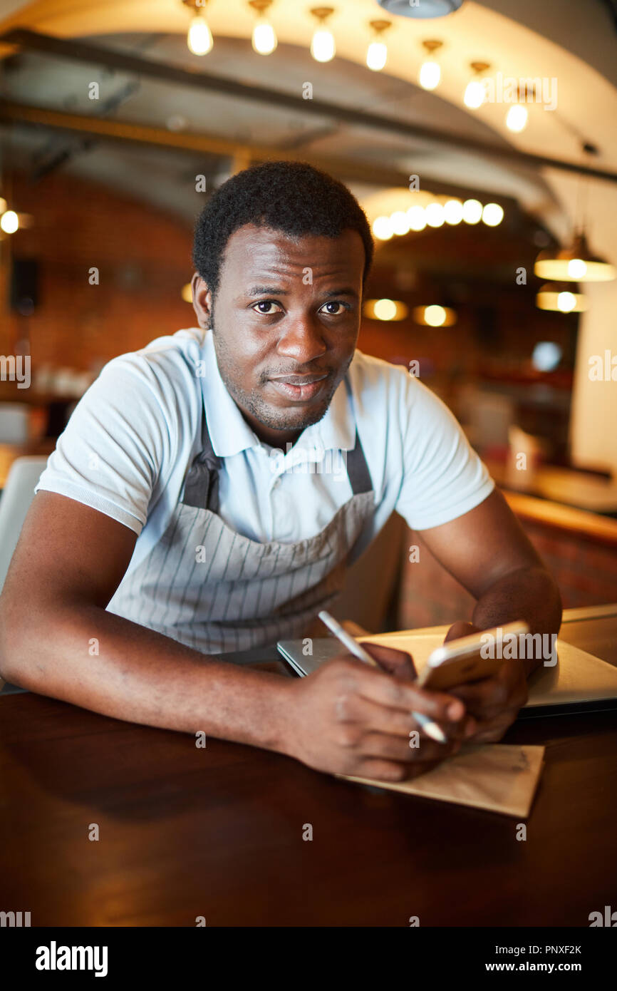 Young waiter or owner of cafe or restaurant in workwear sitting by ...