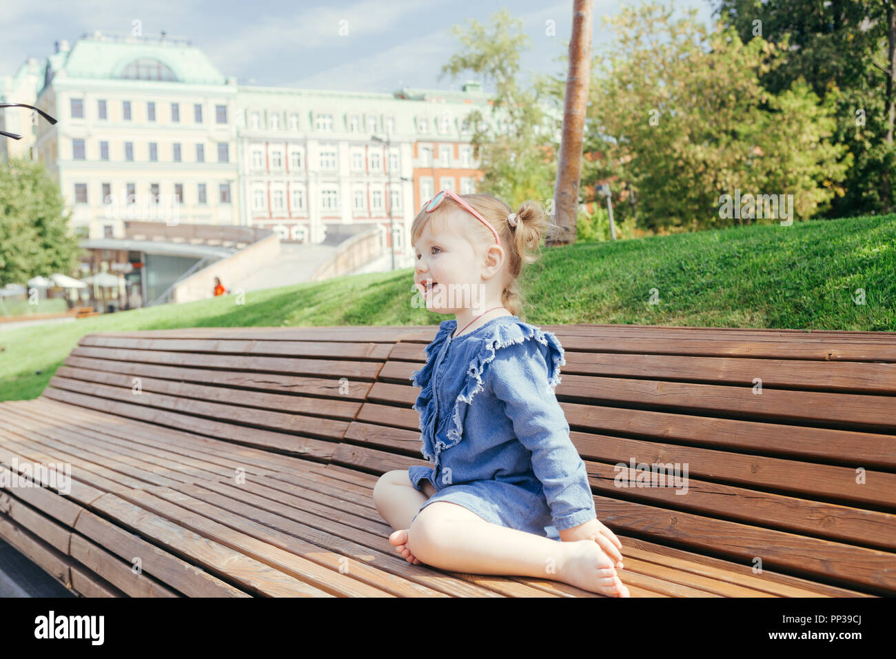 Little girl sitting on the bench in a city park on a warm sunny day ...