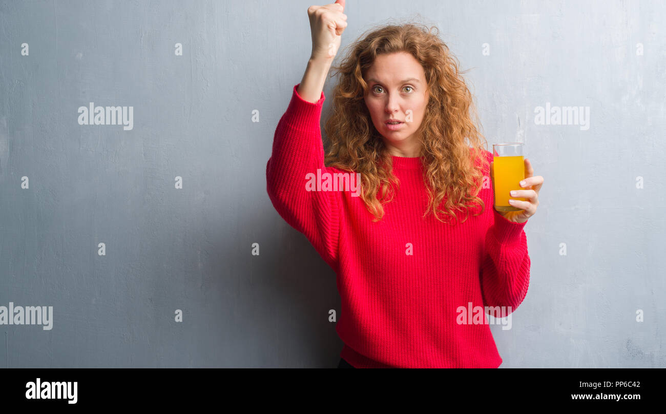 Young redhead woman over grey grunge wall drinking a glass of orange ...