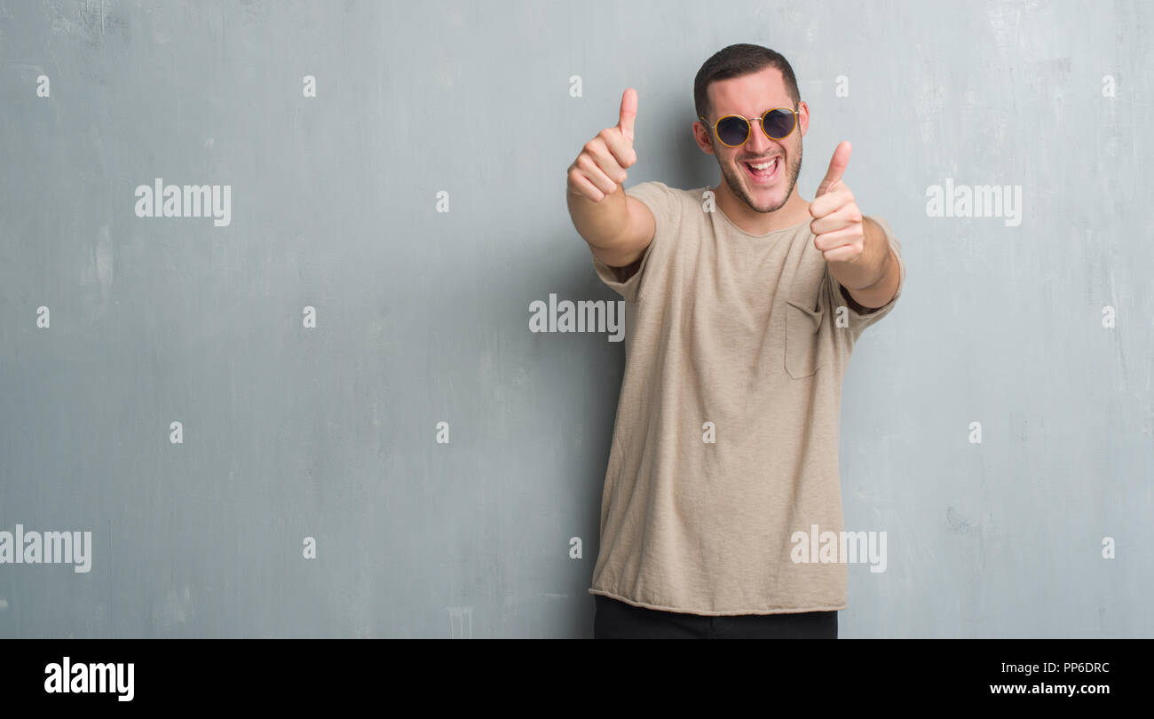 Young caucasian man over grey grunge wall wearing sunglasses approving ...