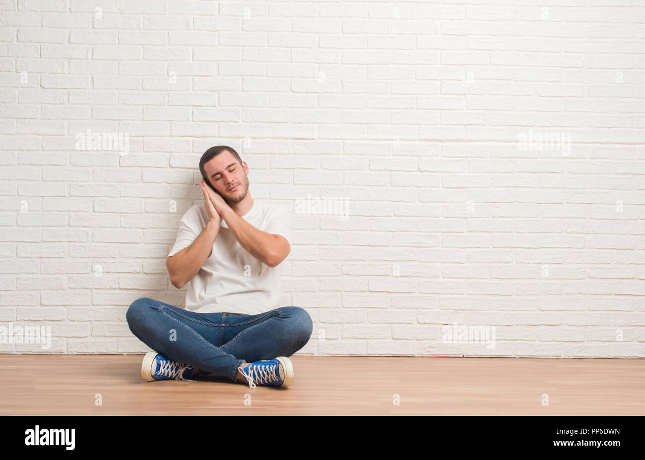Young caucasian man sitting on the floor over white brick wall sleeping ...