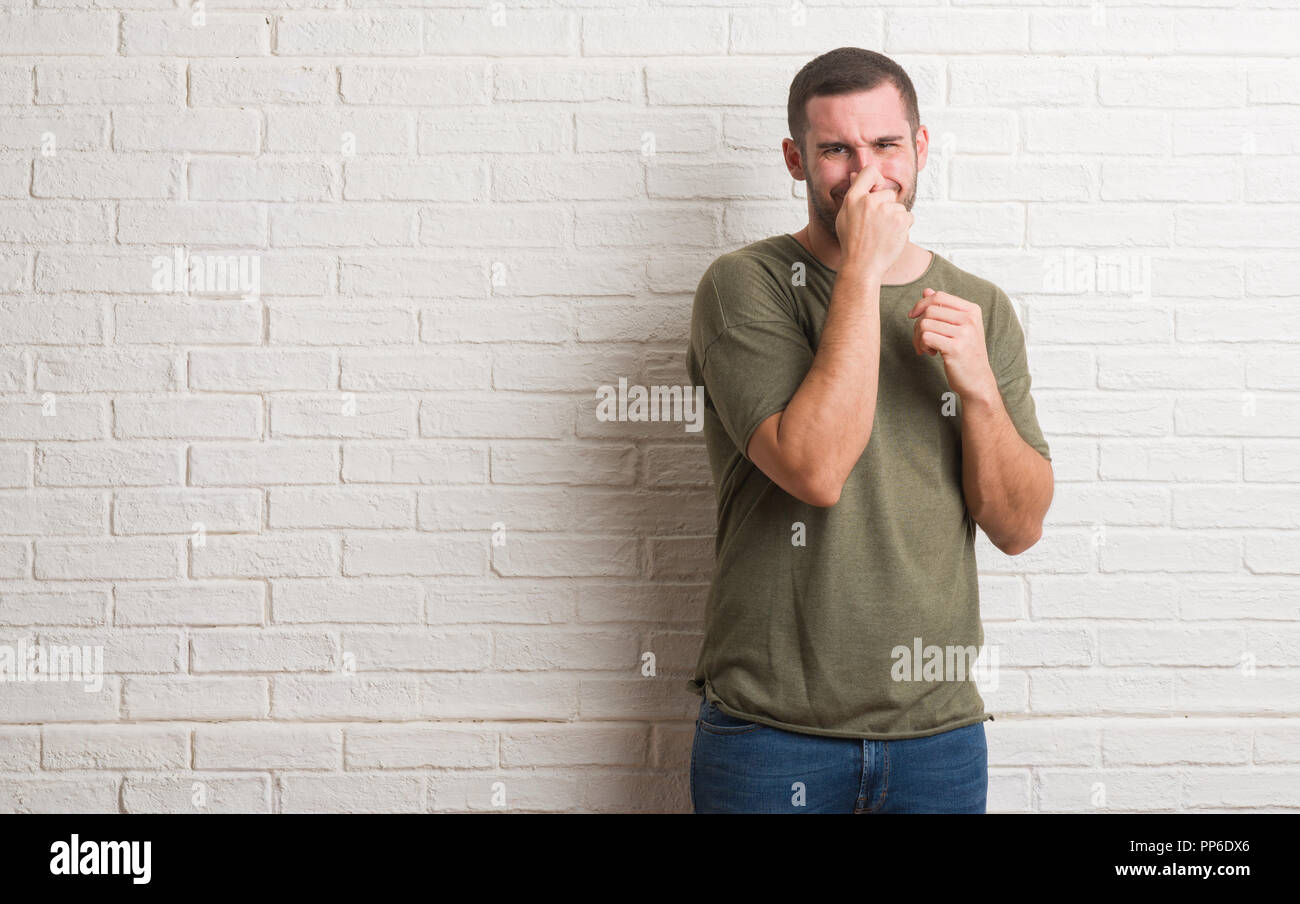 Young caucasian man standing over white brick wall smelling something ...