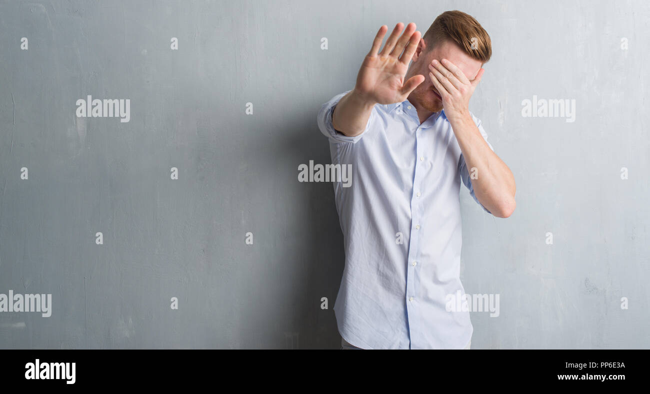 Young redhead business man over grey grunge wall covering eyes with ...