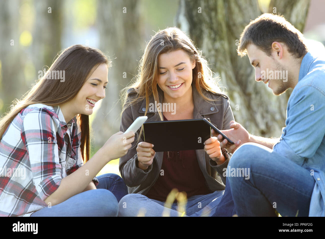 Three happy friends using multiple devices sitting on the grass in a ...