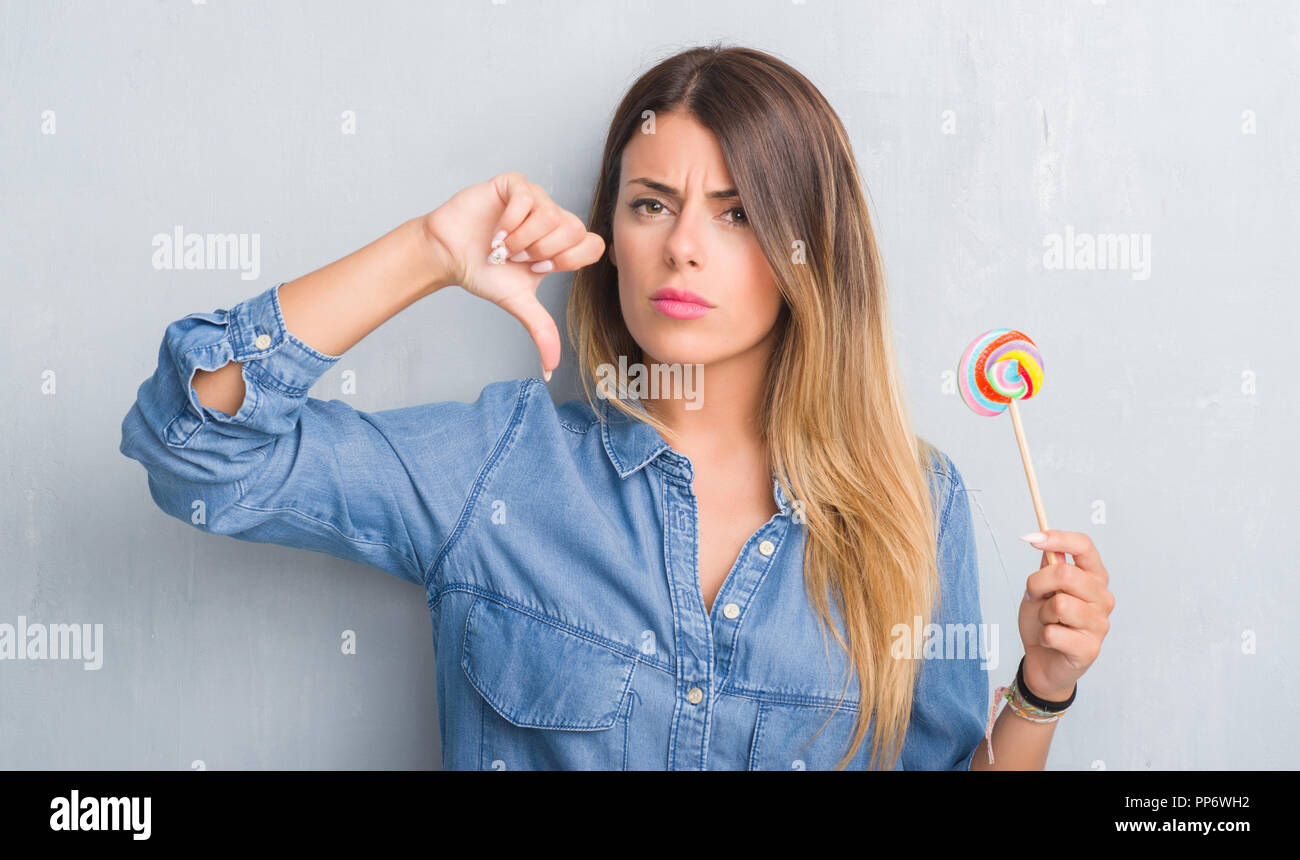 Young adult woman over grey grunge wall eating lollipop candy with ...