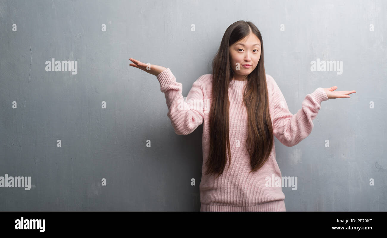 Young Chinese woman over grey wall clueless and confused expression ...