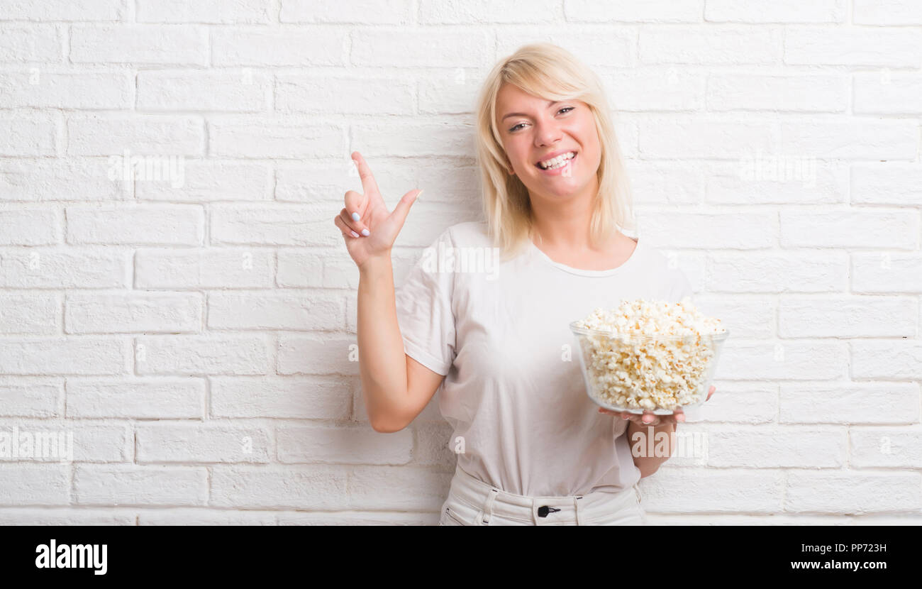 Adult caucasian woman over white brick wall eating pop corn very happy ...