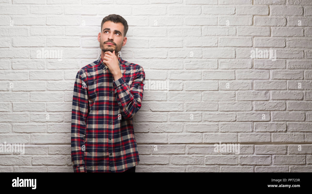 Young adult man over white brick wall serious face thinking about ...