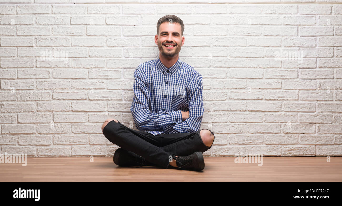 Young adult man sitting over white brick wall happy face smiling with ...
