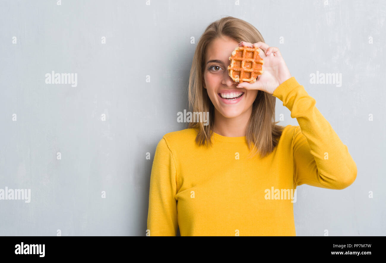 Beautiful young woman over grunge grey wall eating waffle with a happy ...