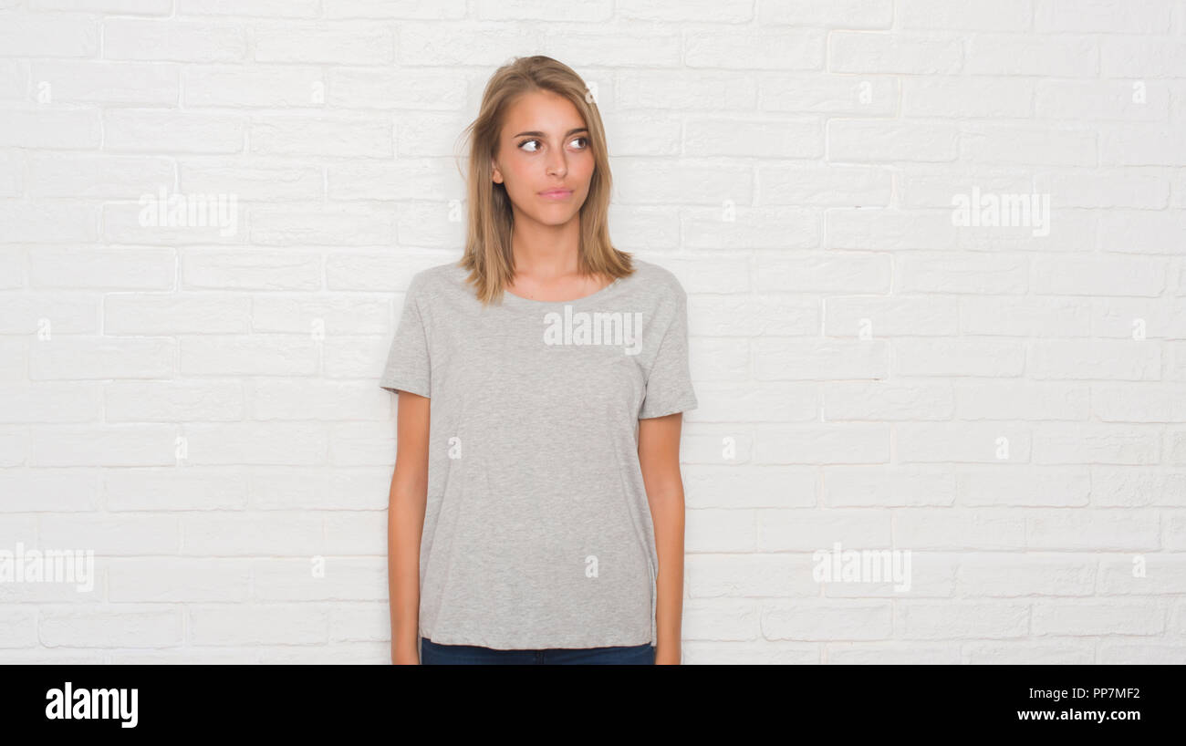 Beautiful young woman over white brick wall smiling looking side and ...