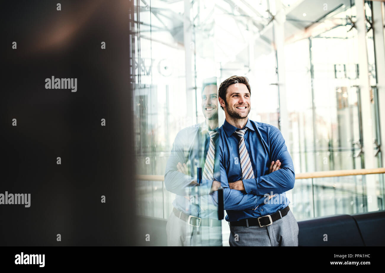 A portrait of a young businessman standing inside a building, arms ...