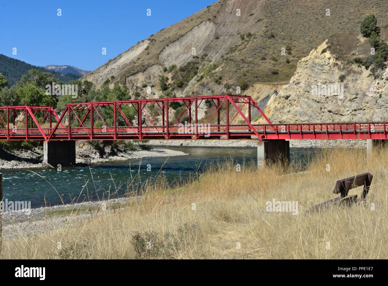 A wooden bench in high grass with a bright red bridge over a mountain river in background Stock ...