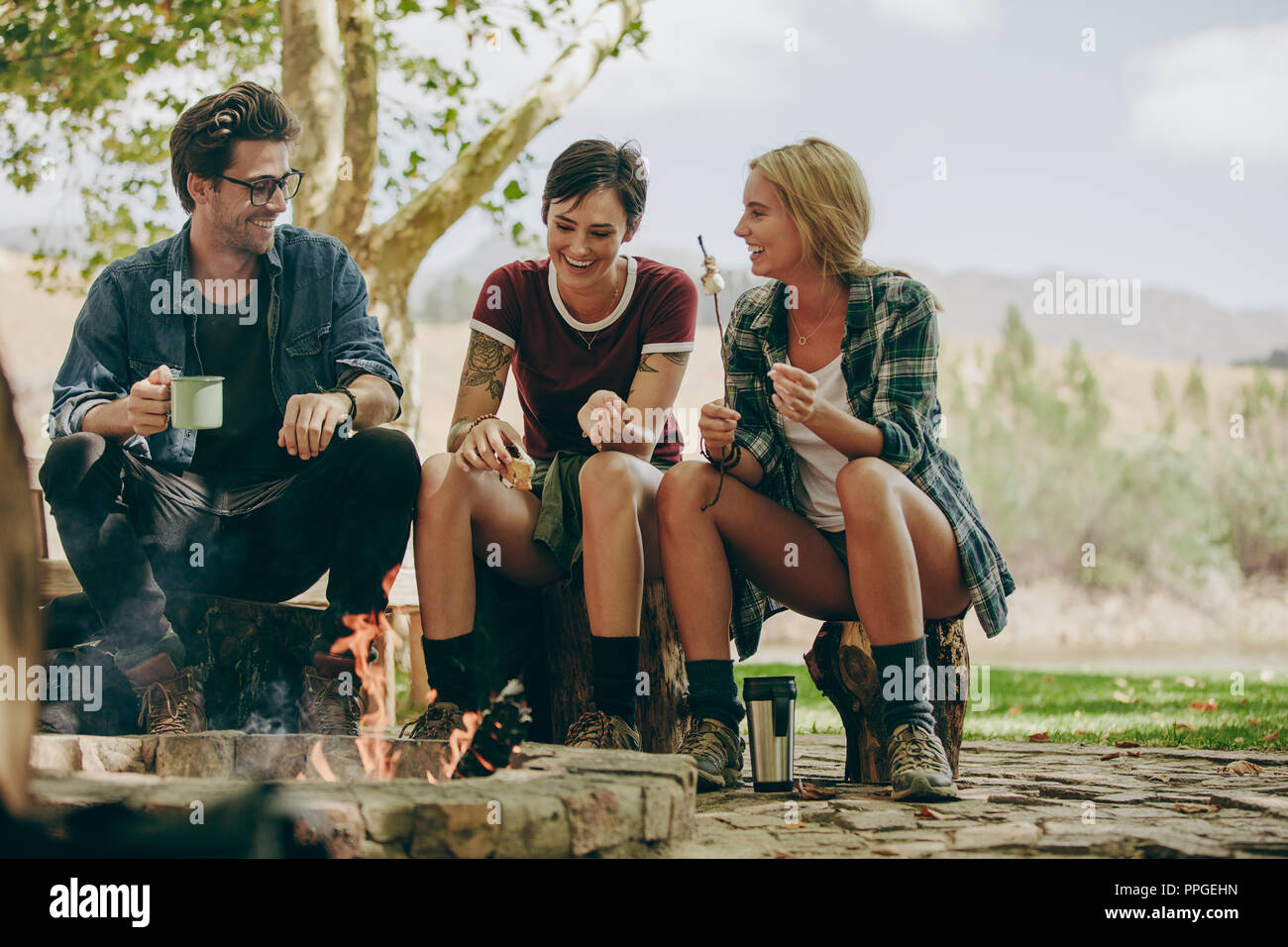 Happy friends on holiday toasting food on bonfire sitting together ...