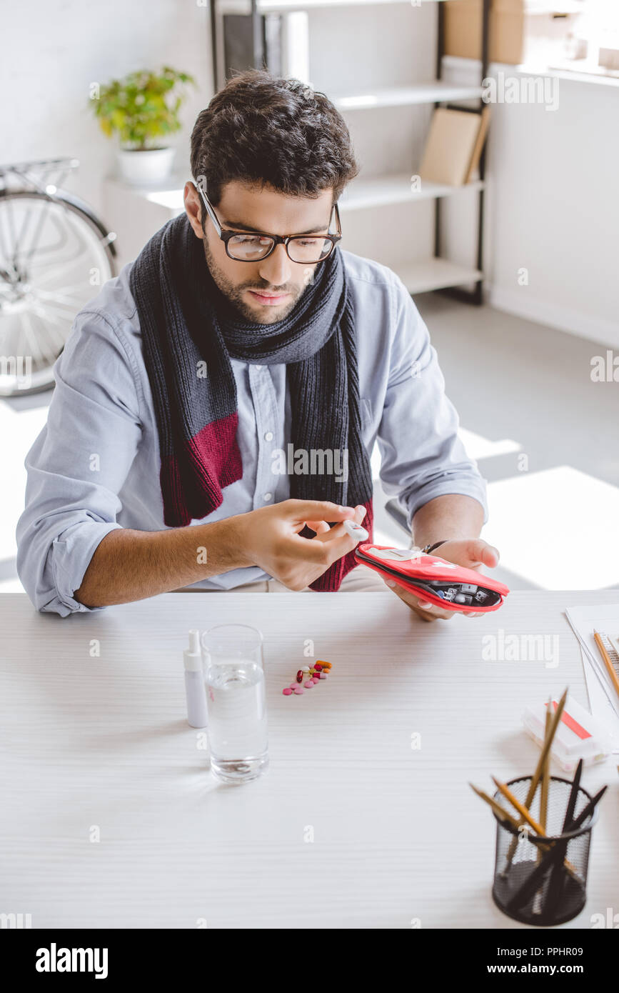 sick manager in scarf holding first aid kit and electronic thermometer ...