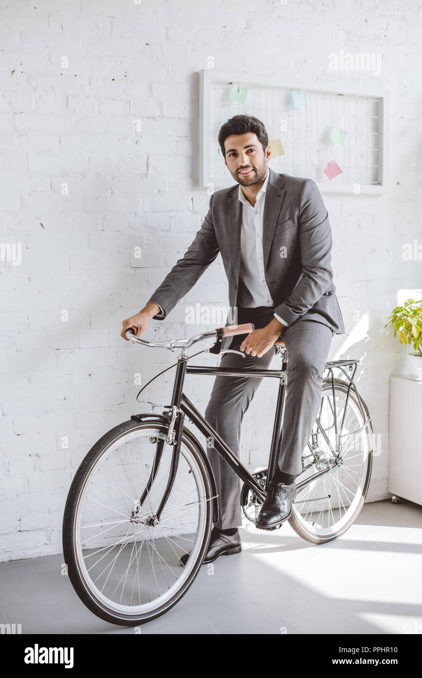smiling businessman sitting on bicycle in office and looking at camera ...
