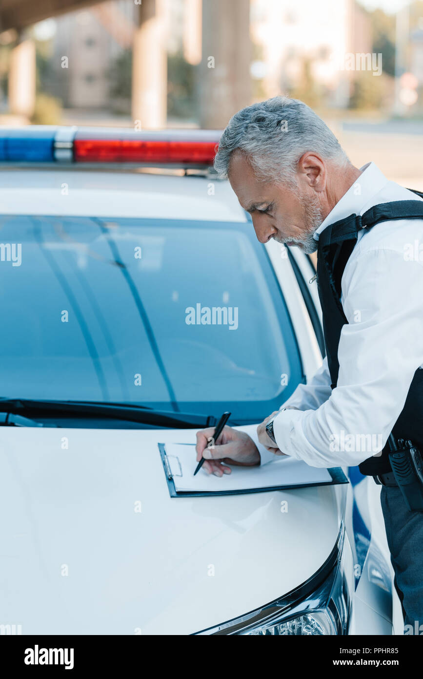 side view of focused male police officer writing in clipboard on car at ...