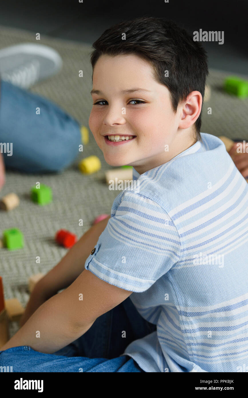 side view of smiling boy looking at camera while sitting on floor with ...