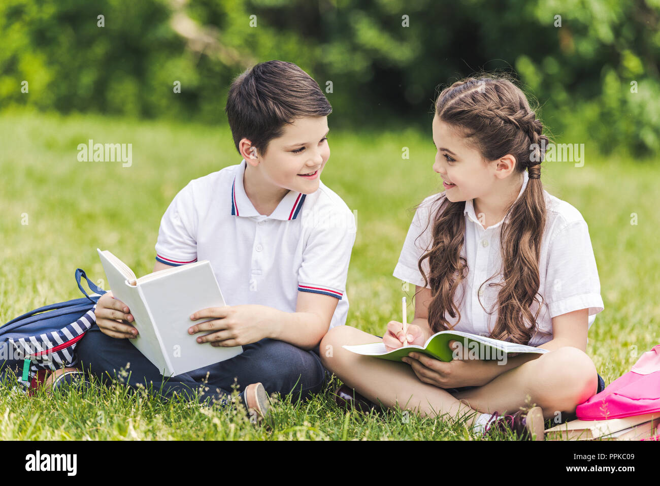 happy schoolchildren doing homework together while sitting on grass in ...