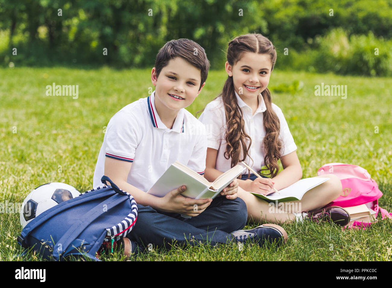 smiling schoolchildren doing homework together while sitting on grass ...
