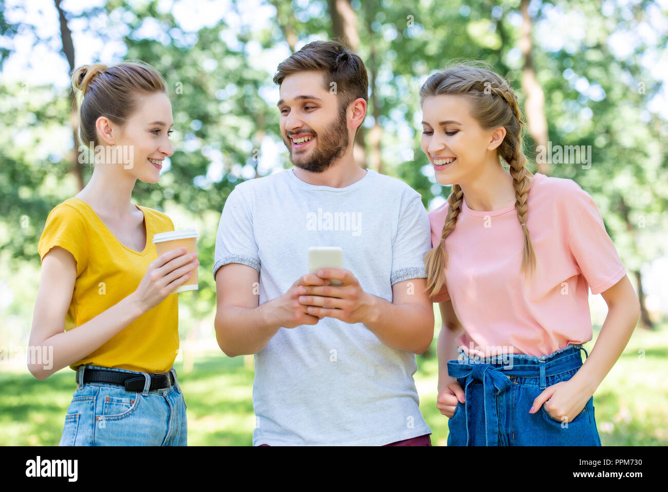 smiling friends with coffee to go using smartphone together in park ...