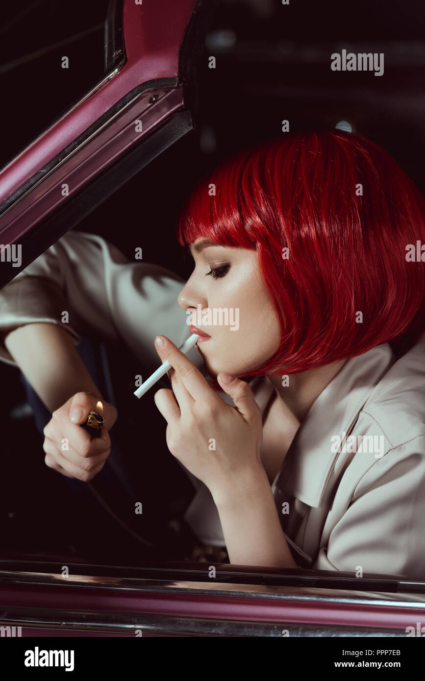 side view of beautiful young woman in red wig smoking cigarette in car ...